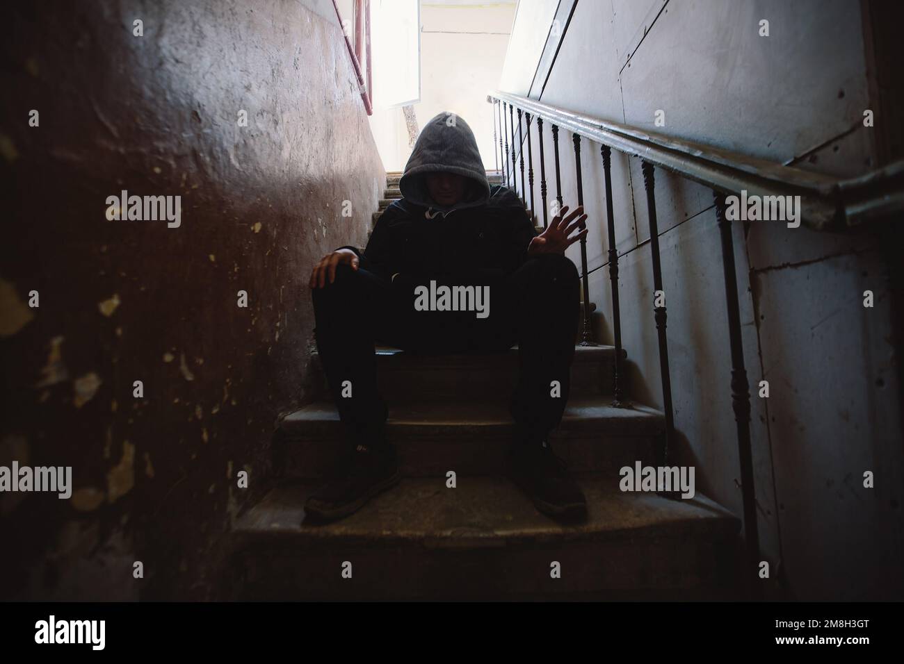 Depressive sad lonely man in hood sits on stairs in shabby house ...