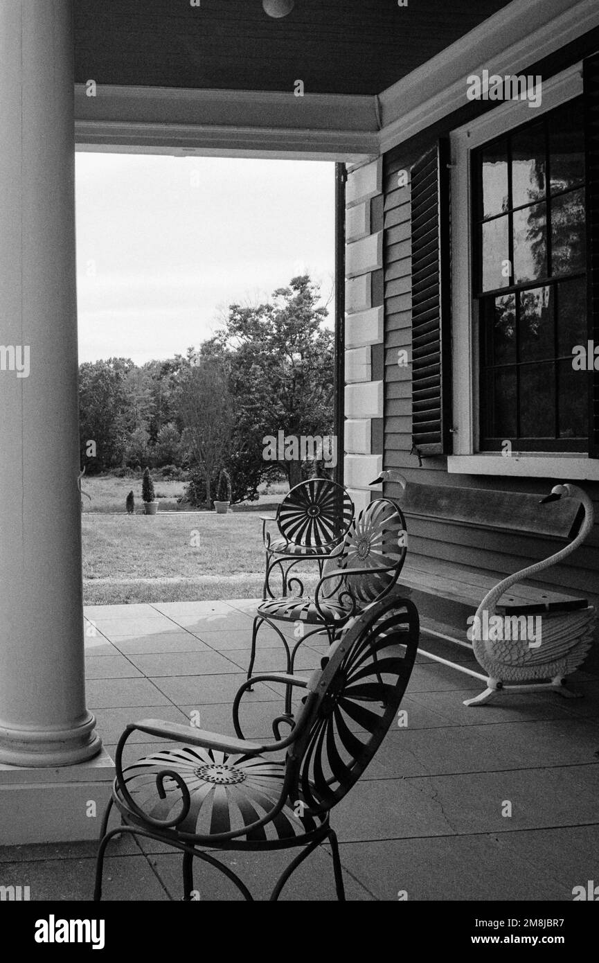 Three Metal Outdoor Chairs And Swan Bench Sit Empty Under A Porch Roof On A Summer Day At The
