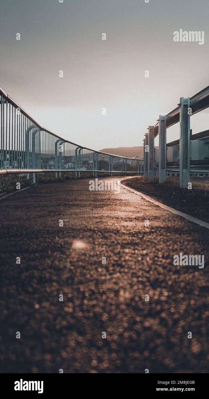 A vertical ground-level shot of an asphalt road with a metallic fence ...