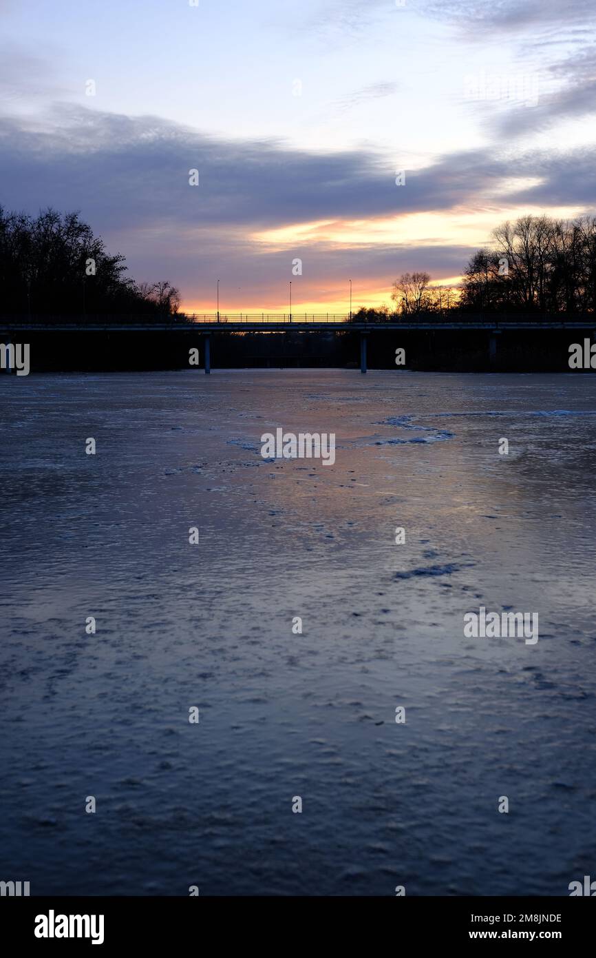 Frosty pink sunset by the frozen river with a bridge going over the water and people walking on ...