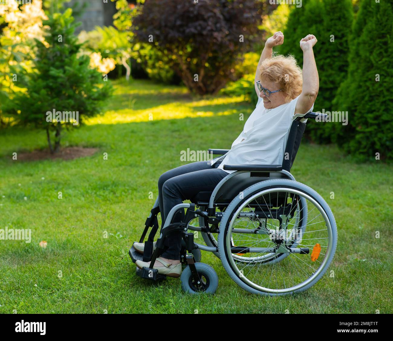 Elderly woman stretches her arms up while sitting in a wheelchair on a ...