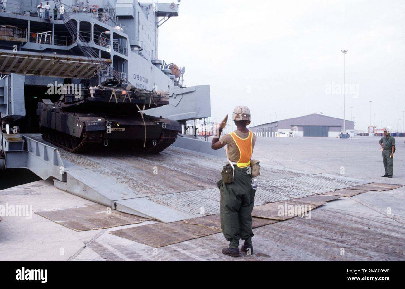 A US Army stevedore directs a M-1 Abrams Main Battle Tank down the ramp ...