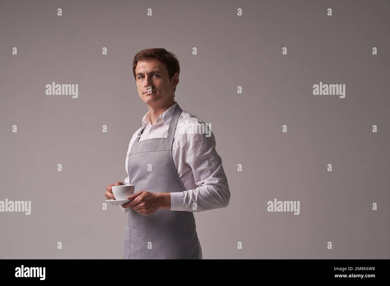 A handsome young smiling man in grey apron standing alone against a ...