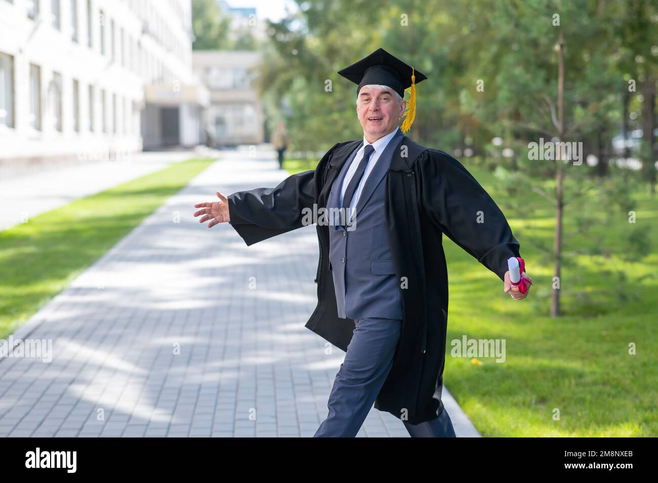 An old man in a graduation gown walks outdoors and holds a diploma ...