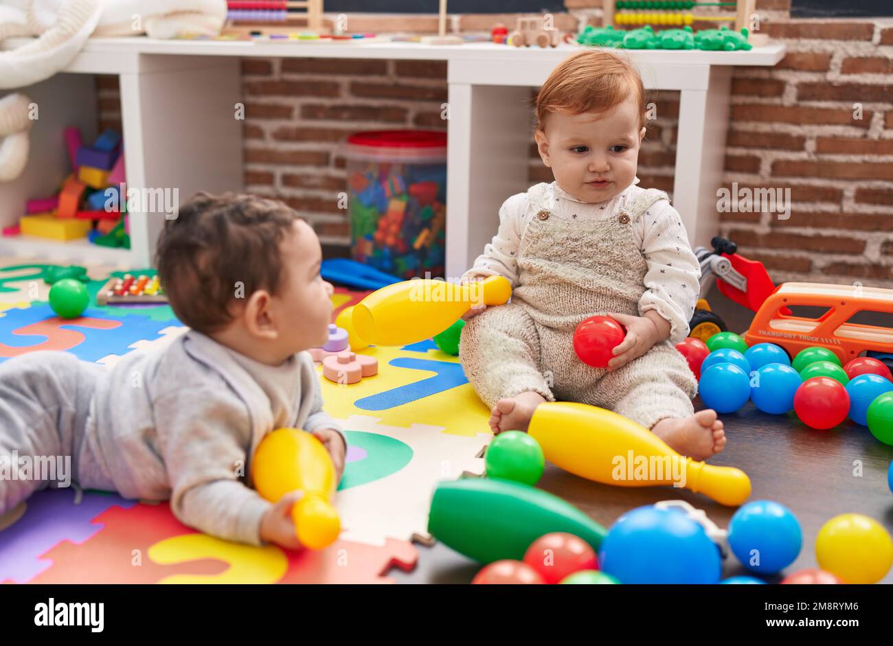 Two adorable babies playing with balls and bowling pin sitting on floor ...