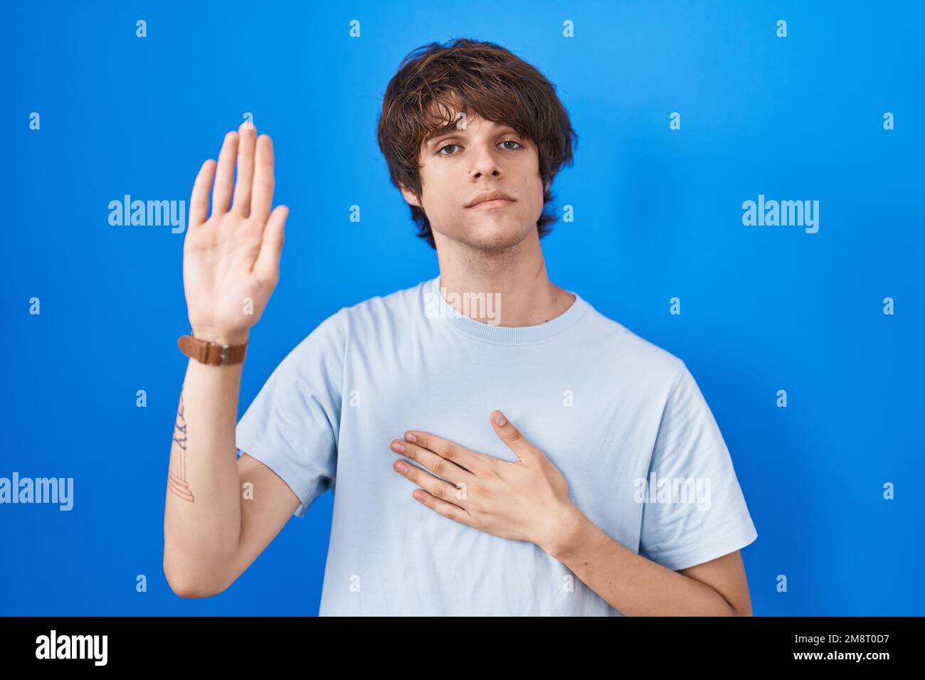 Hispanic young man standing over blue background swearing with hand on ...