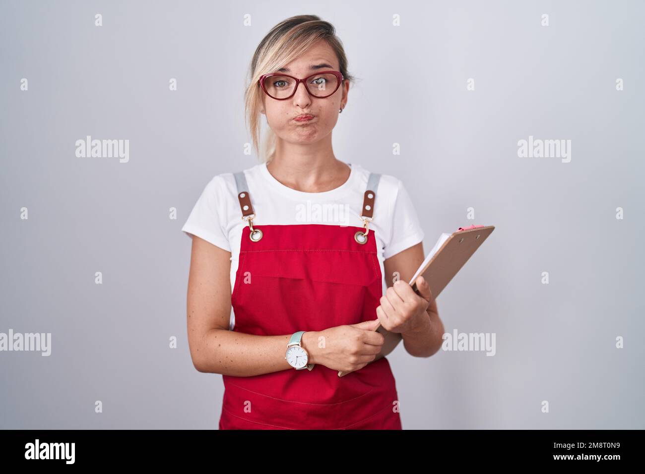 Young blonde woman wearing waiter uniform holding clipboard puffing ...