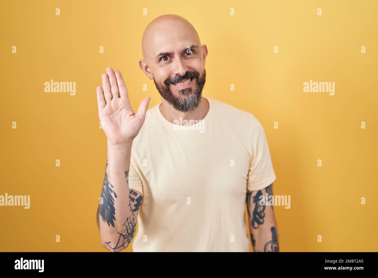 Hispanic man with tattoos standing over yellow background waiving ...