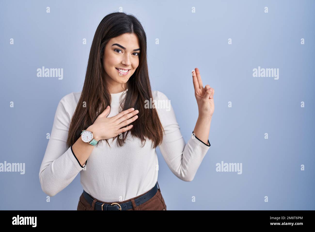 Young brunette woman standing over blue background smiling swearing ...