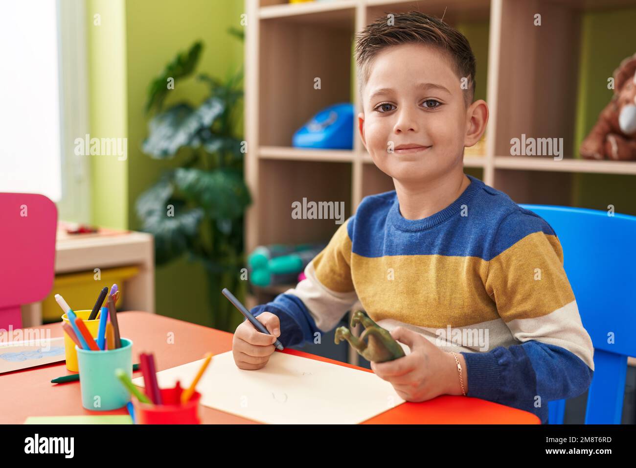 Adorable hispanic boy student sitting on table drawing on paper at ...