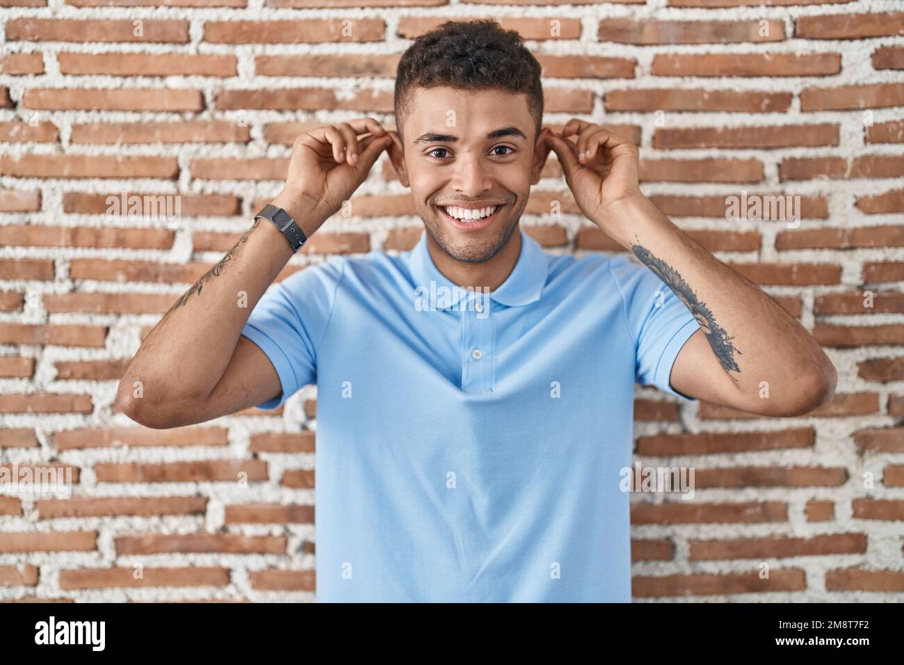 Brazilian young man standing over brick wall smiling pulling ears with ...