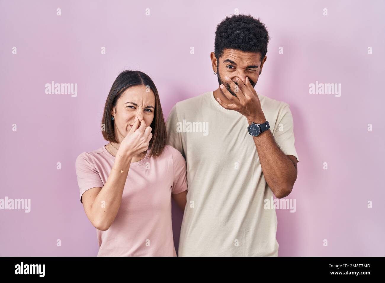 Young hispanic couple together over pink background smelling something ...