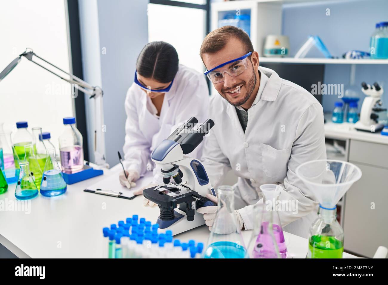 Man and woman scientist partners working using microscope and writing on clipboard at laboratory ...