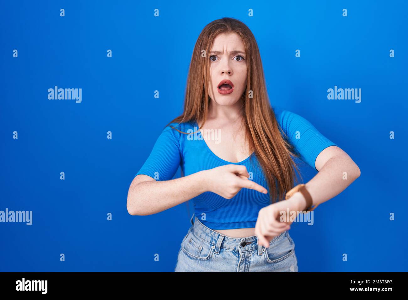 Redhead woman standing over blue background in hurry pointing to watch ...