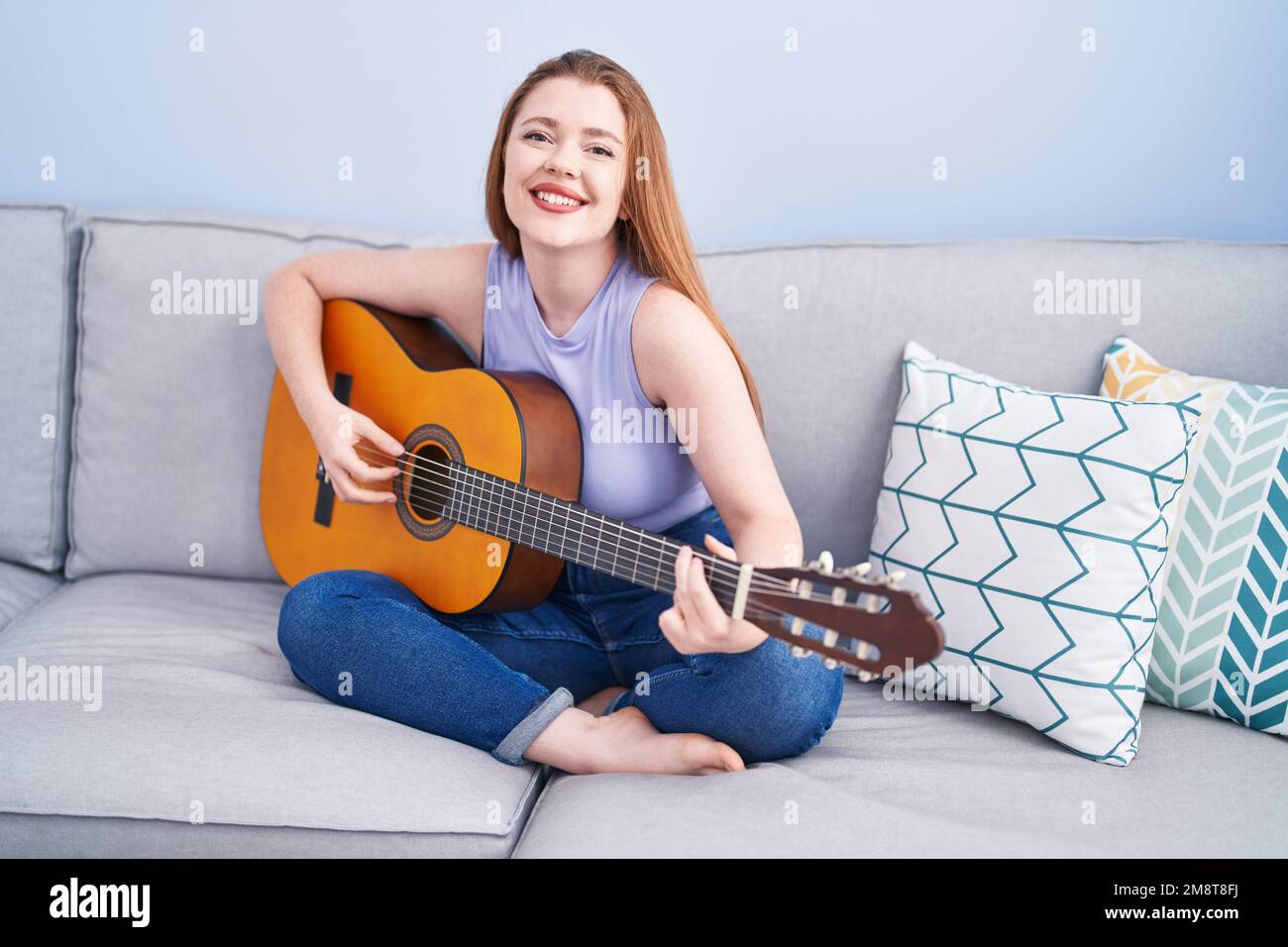 Young redhead woman playing classical guitar sitting on sofa at home ...