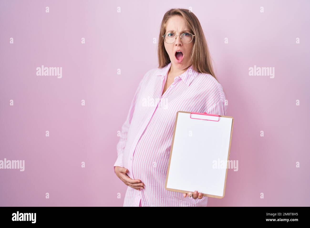 Young pregnant woman holding clipboard afraid and shocked with surprise ...