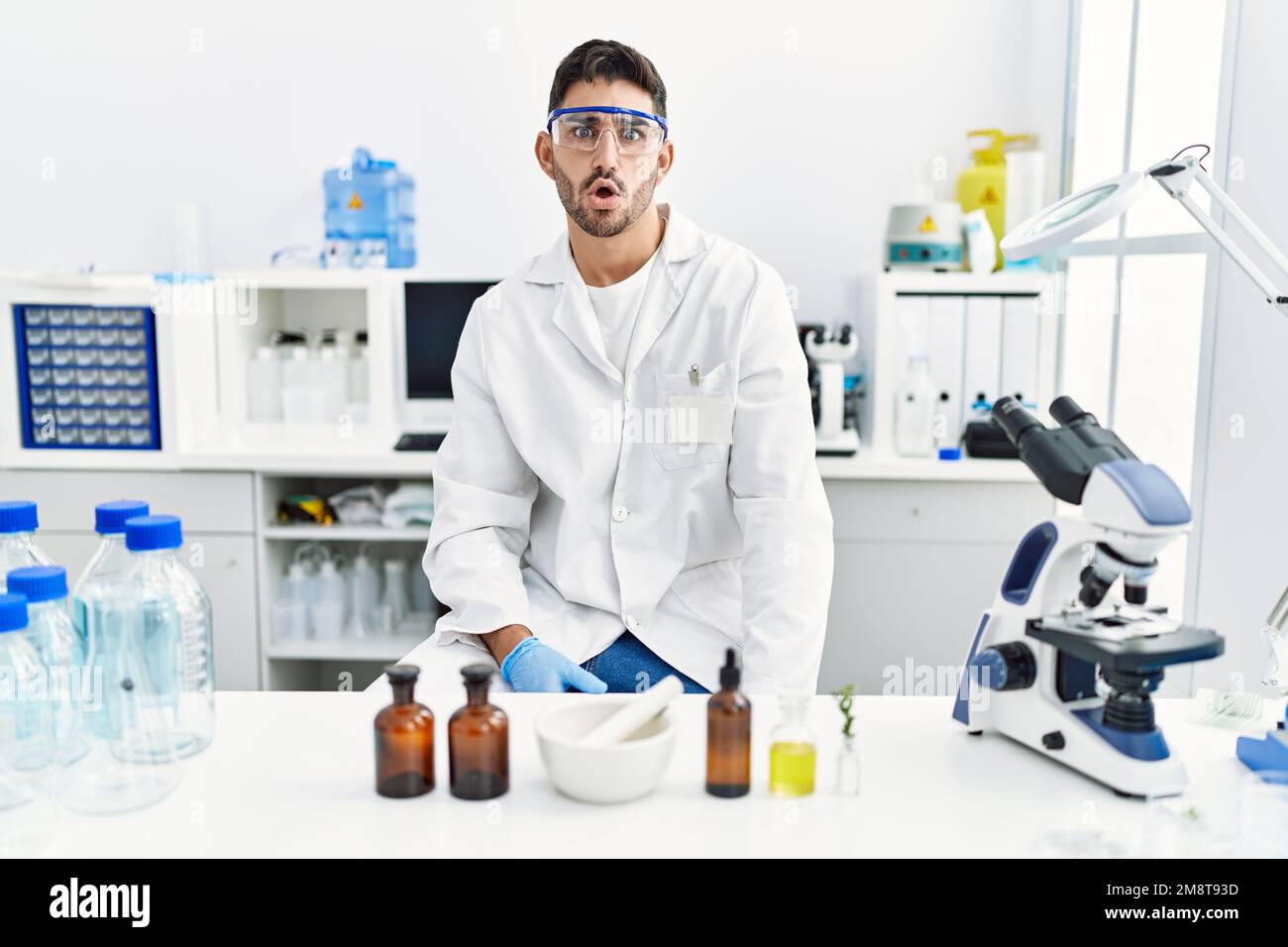 Young hispanic man working at scientist laboratory scared and amazed ...