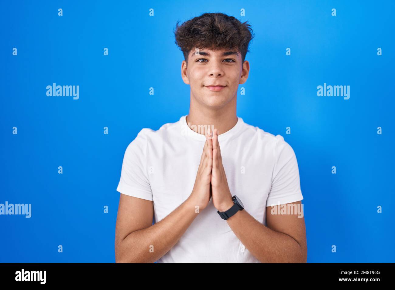 Hispanic teenager standing over blue background praying with hands ...