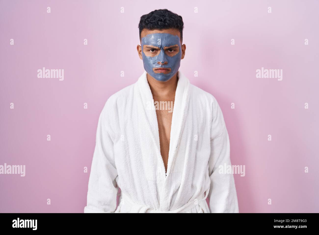 Young hispanic man wearing beauty face mask and bath robe skeptic and ...