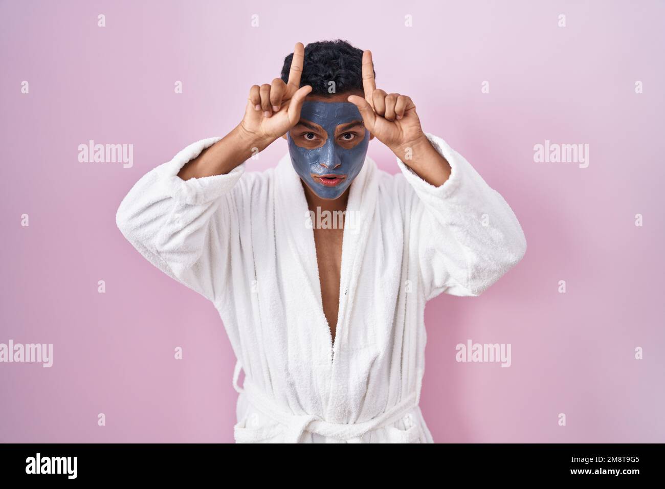 Young hispanic man wearing beauty face mask and bath robe doing funny ...
