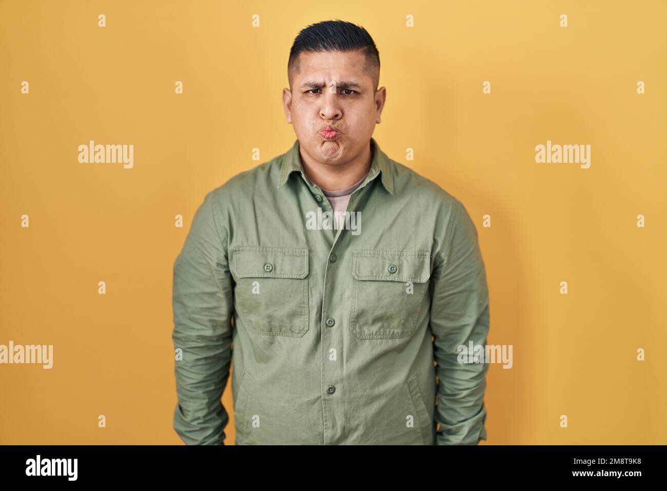 Hispanic young man standing over yellow background puffing cheeks with ...