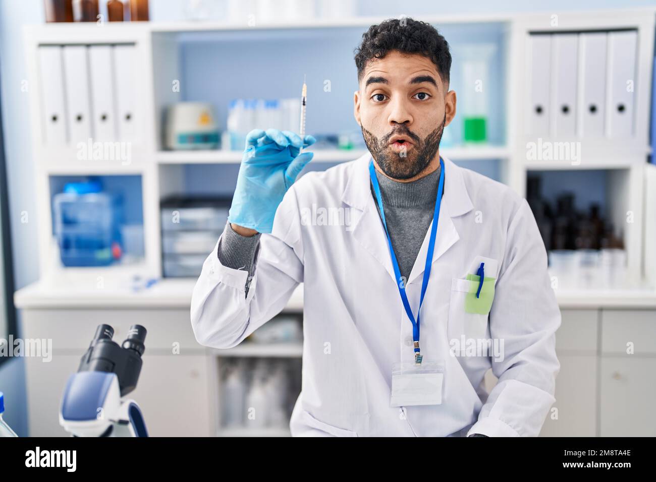 Hispanic man with beard working at scientist laboratory holding syringe scared and amazed with ...