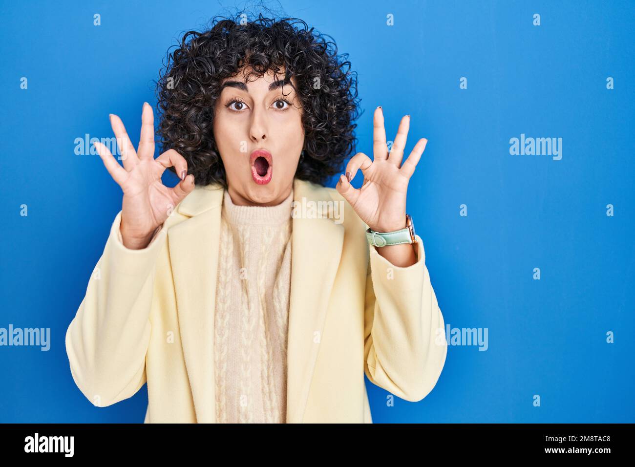 Young brunette woman with curly hair standing over blue background ...