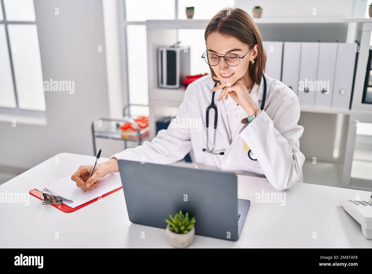 Young caucasian woman doctor using laptop writing on document at clinic ...
