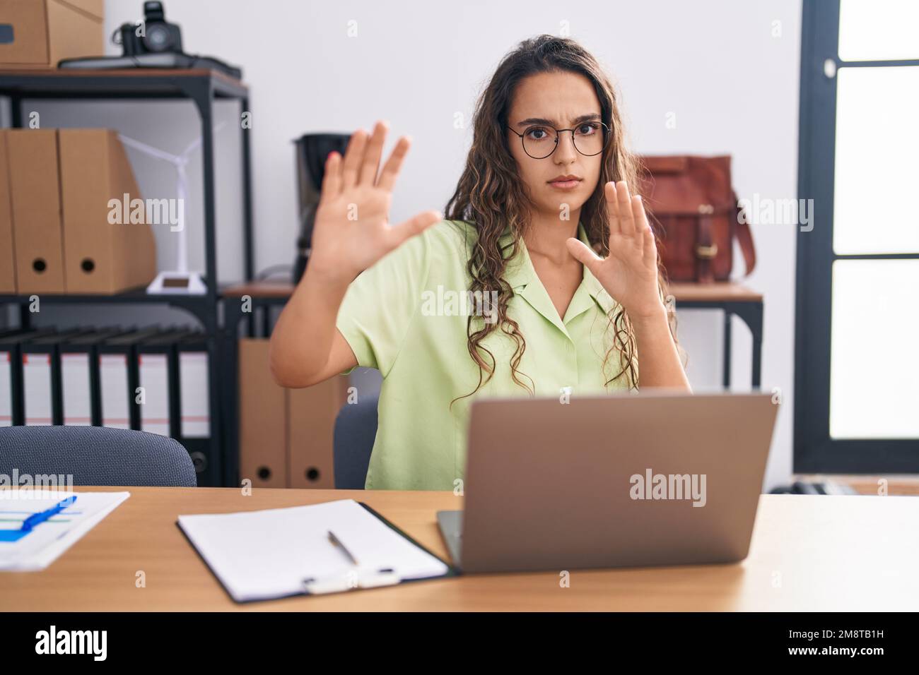 Young hispanic woman working at the office wearing glasses afraid and ...