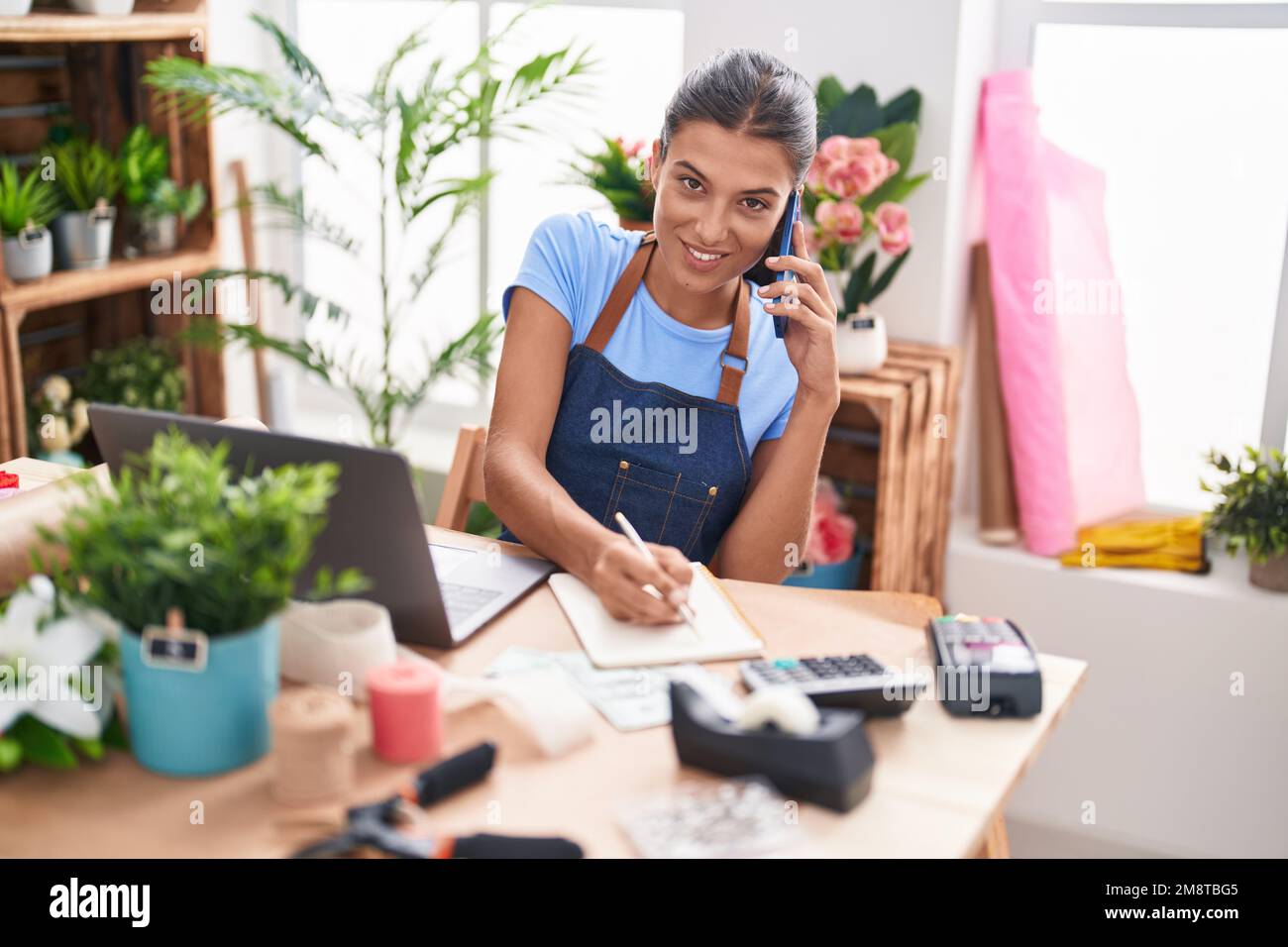 Young beautiful hispanic woman florist talking on smartphone writing on ...
