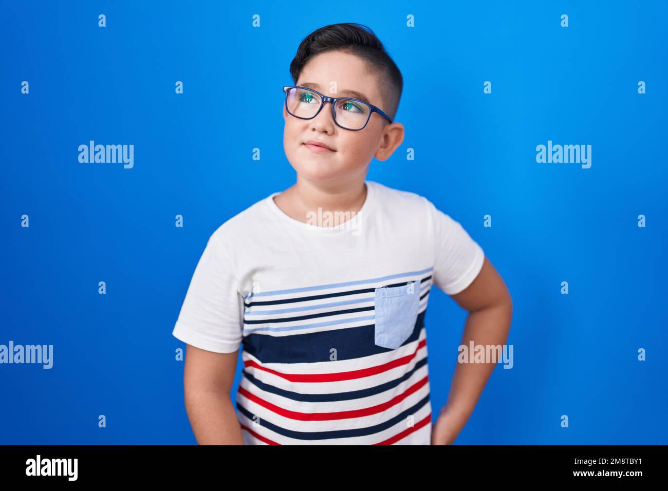 Young hispanic kid standing over blue background smiling looking to the ...