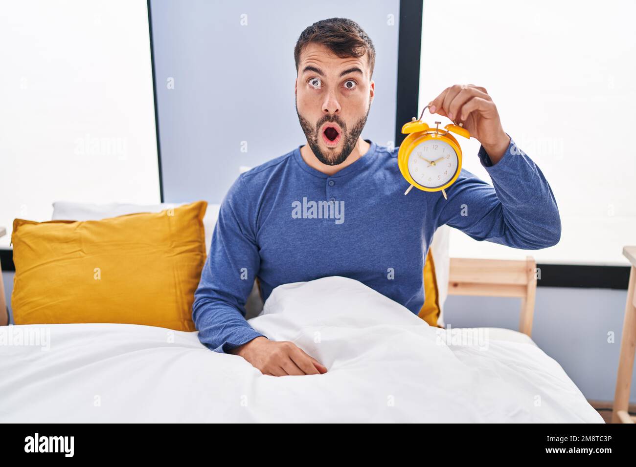 Handsome hispanic man in the bed holding alarm clock scared and amazed ...