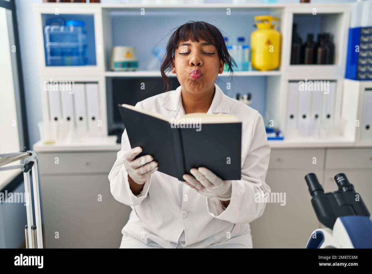 Hispanic woman working at scientist laboratory reading a book puffing ...
