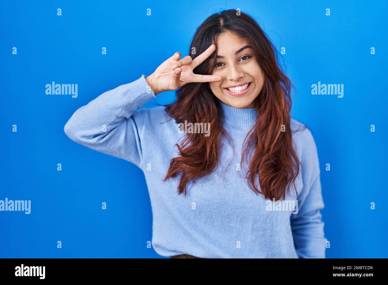 Hispanic young woman standing over blue background doing peace symbol ...