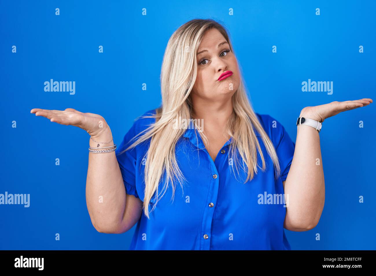 Caucasian plus size woman standing over blue background clueless and ...