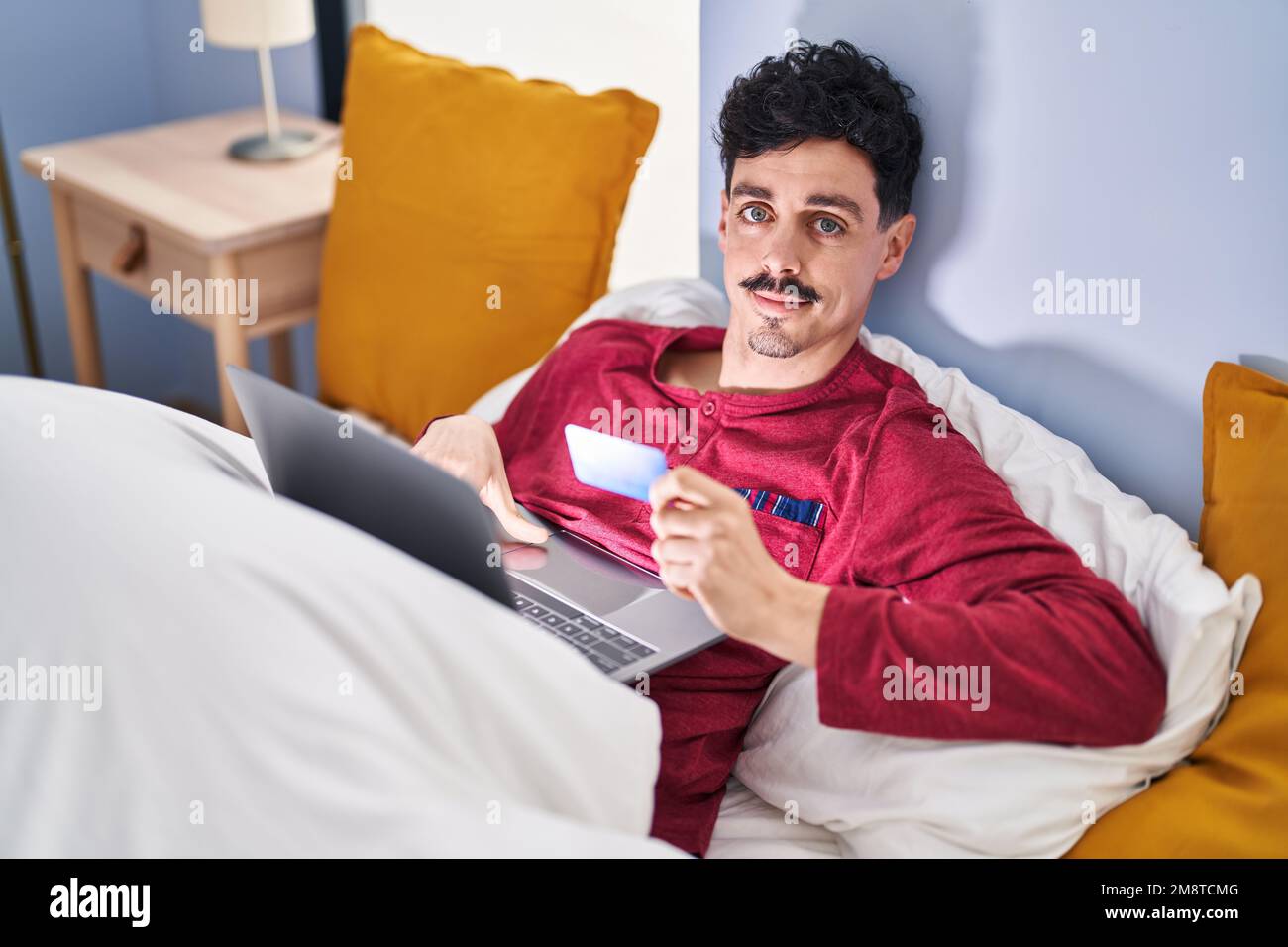 Young caucasian man using laptop and credit card lying on bed at ...