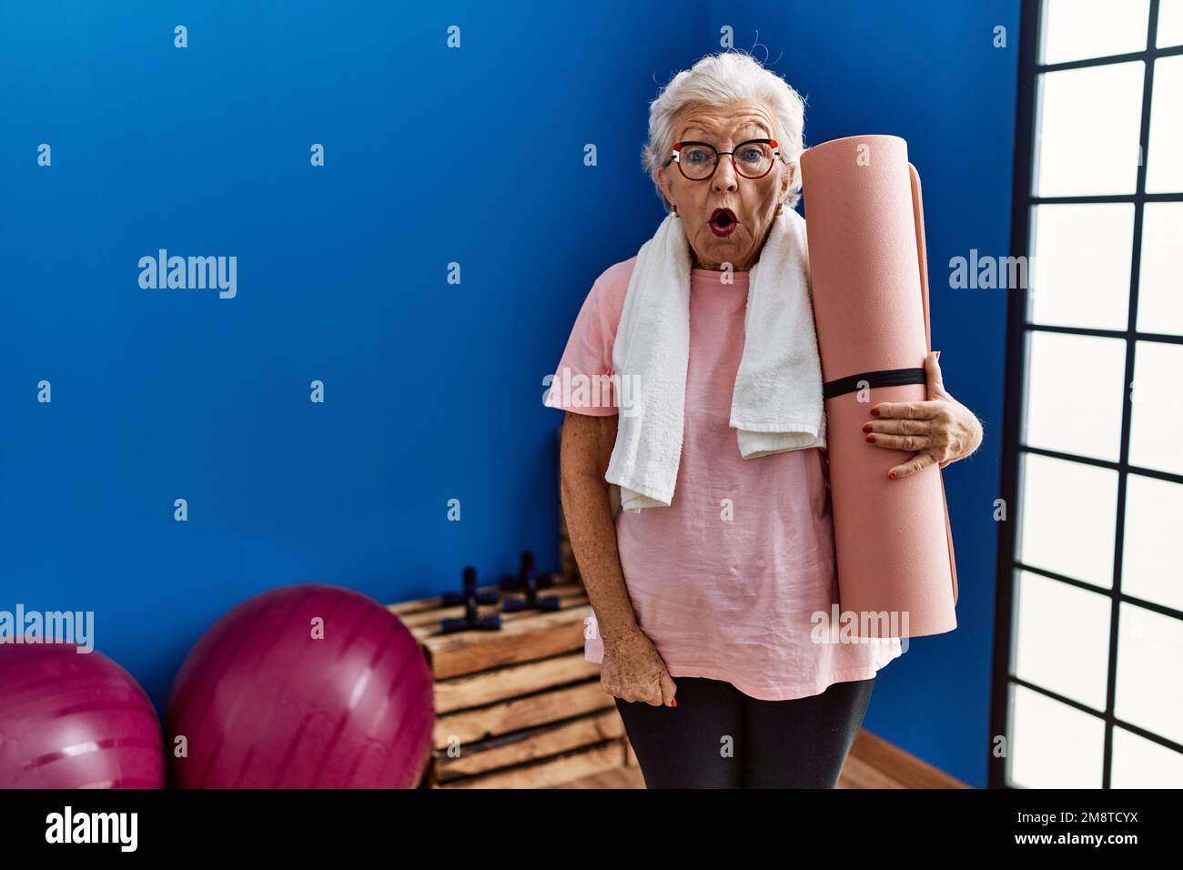 Senior woman with grey hair holding yoga mat scared and amazed with ...