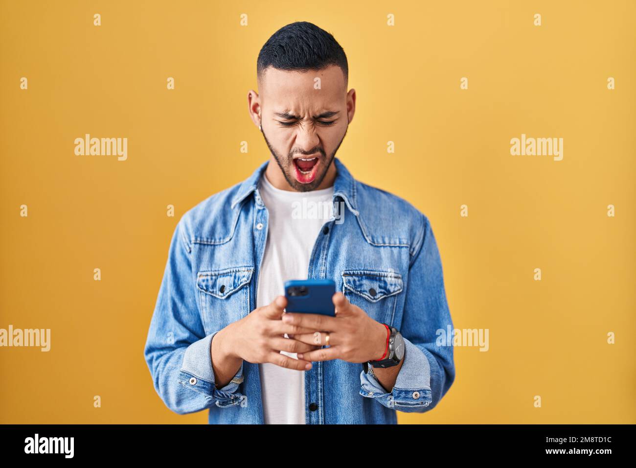 Young hispanic man using smartphone typing message angry and mad ...