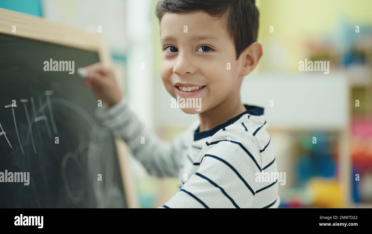 Adorable hispanic boy preschool student writing on blackboard at ...