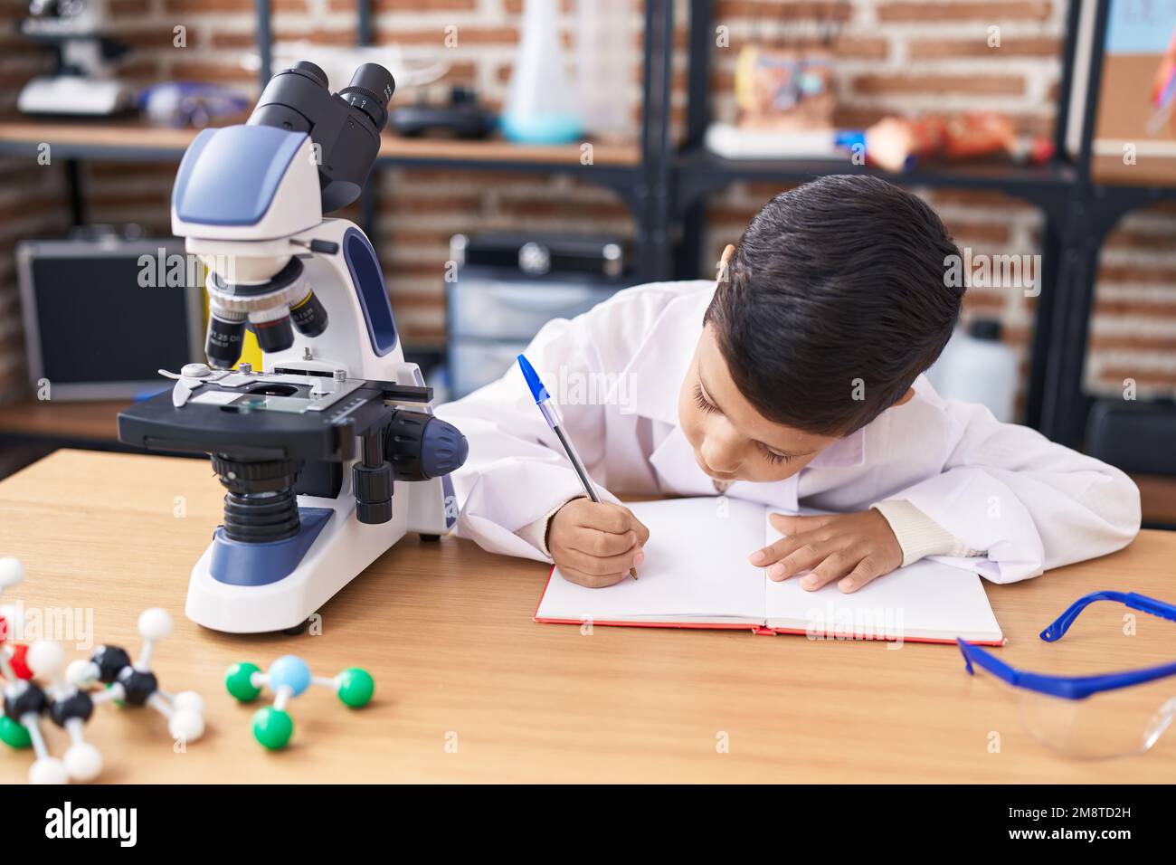 Adorable hispanic boy student using microscope writing on notebook at ...
