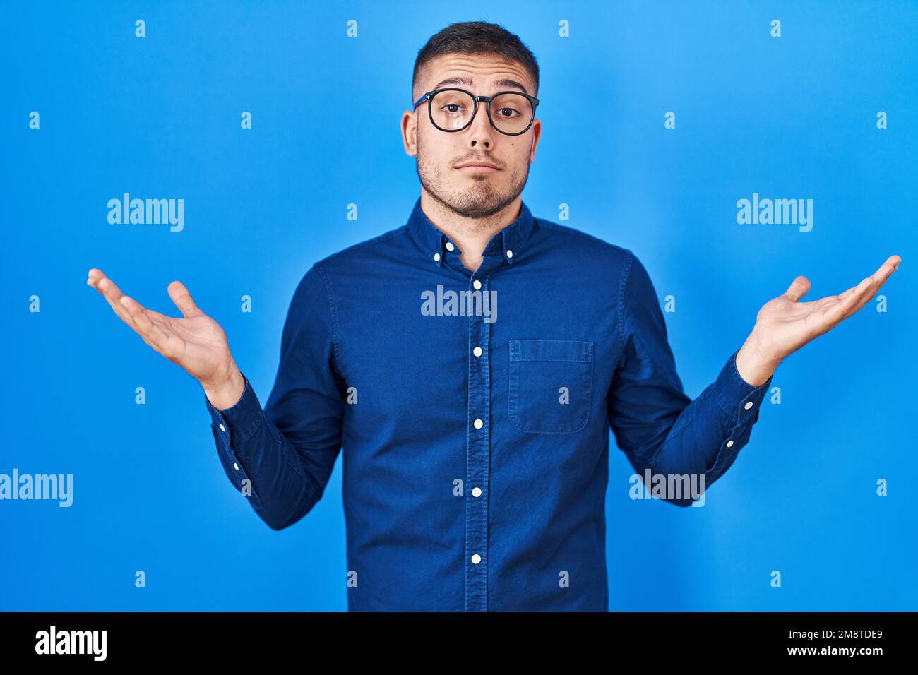 Young hispanic man wearing glasses over blue background clueless and ...