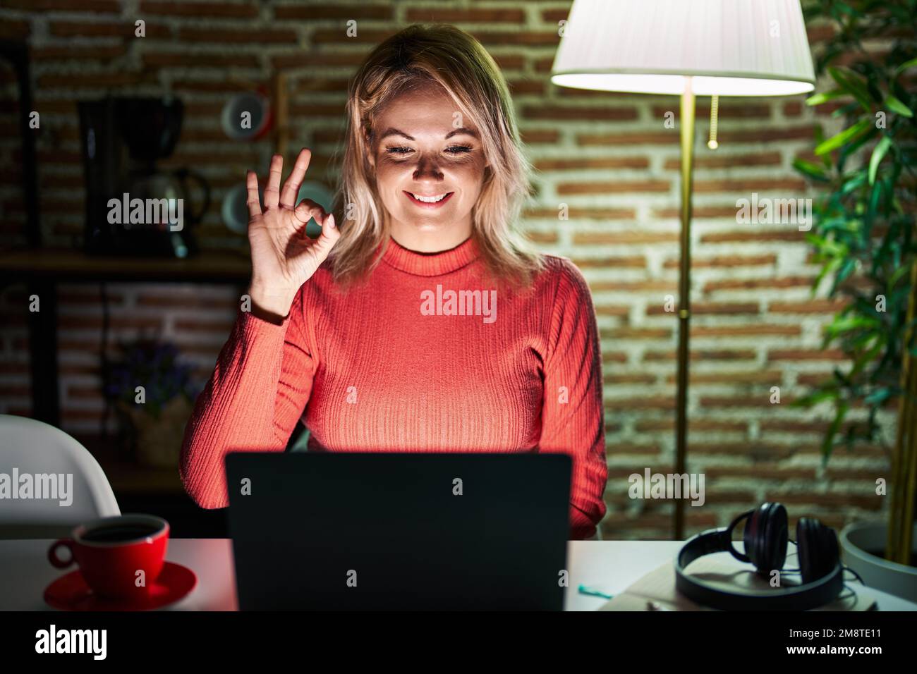Blonde woman using laptop at night at home smiling positive doing ok ...