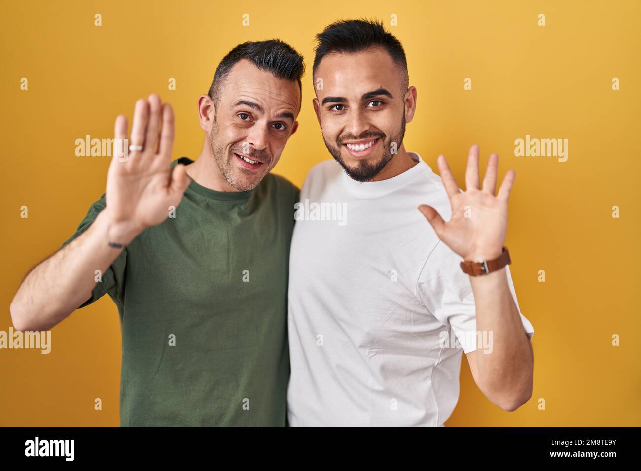 Homosexual couple standing over yellow background waiving saying hello ...