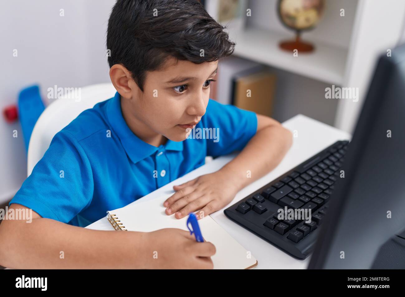 Adorable hispanic boy student using computer writing on notebook at ...