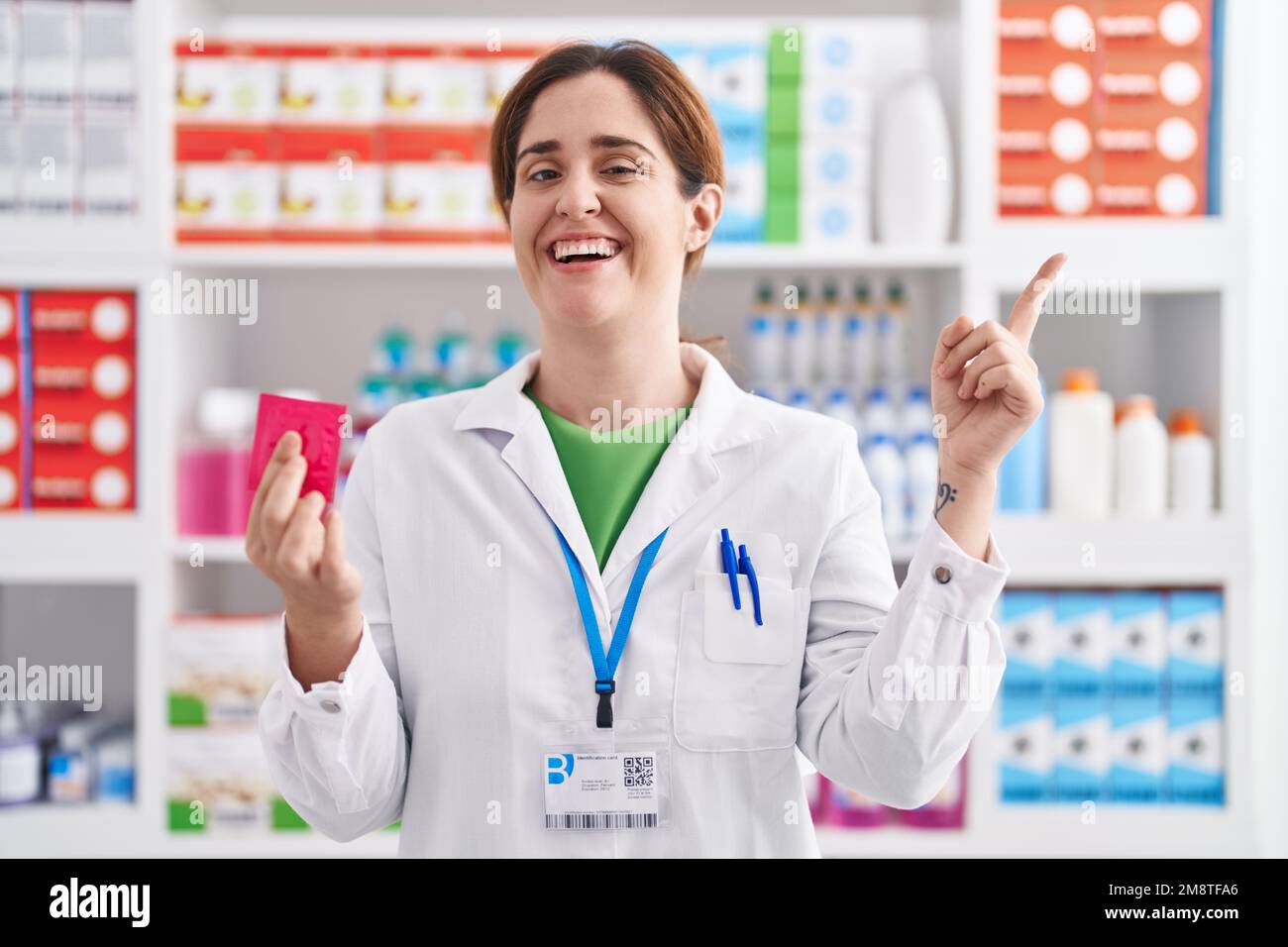 Brunette woman working at pharmacy drugstore holding condom smiling ...