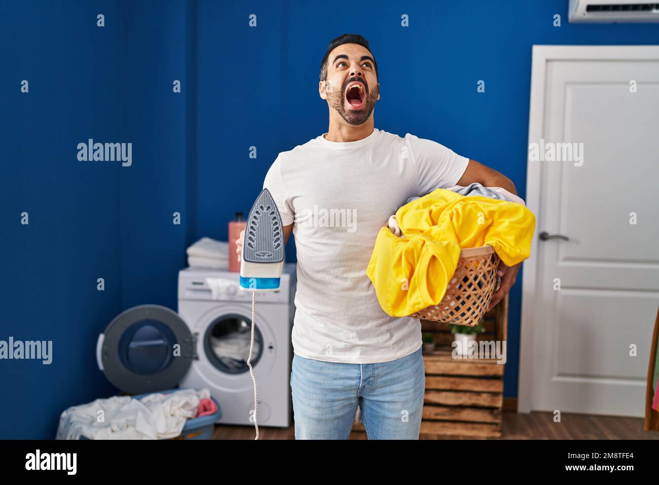 Young hispanic man with beard holding iron and clothes at home angry ...