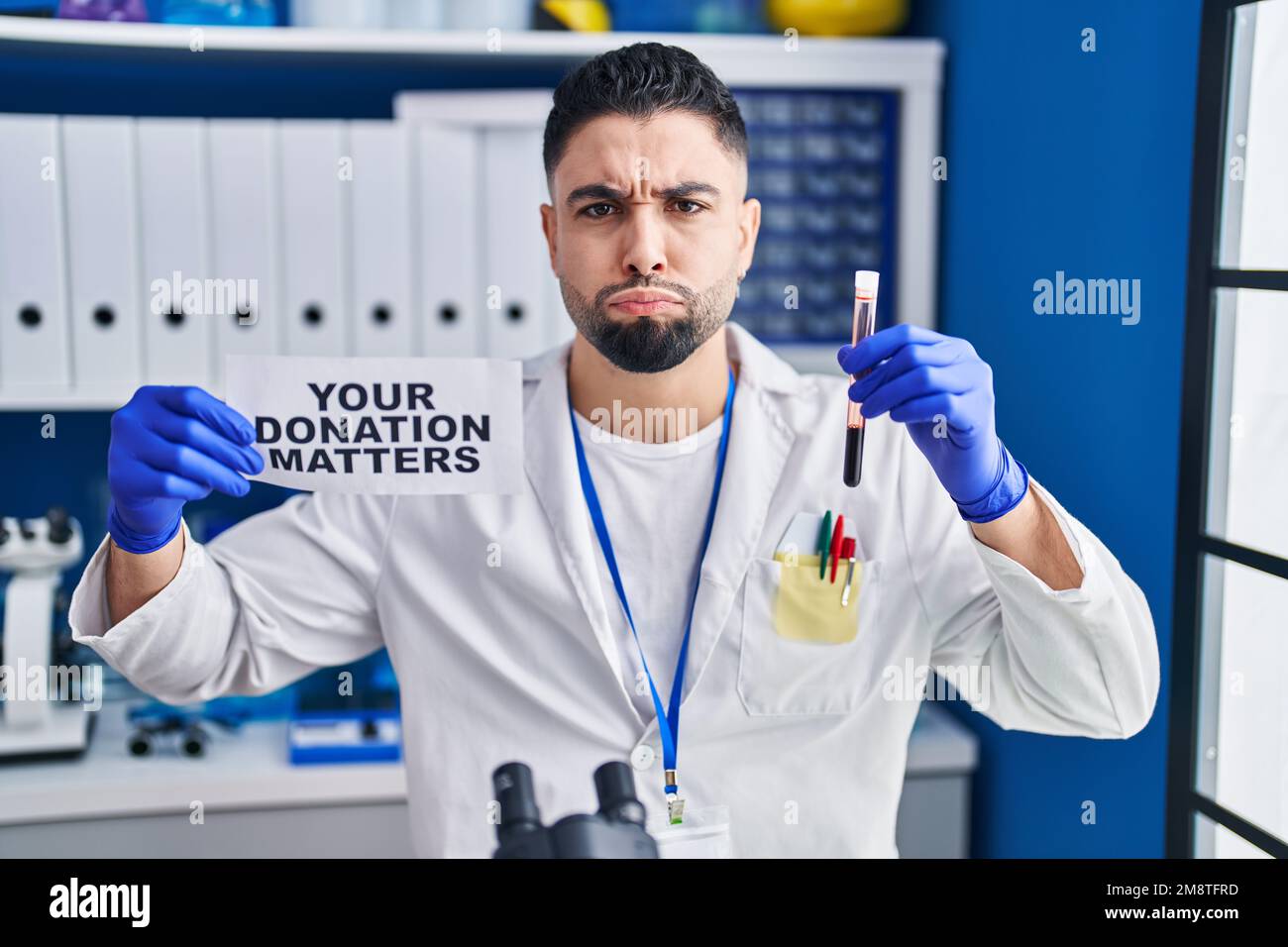 Young handsome man working at scientist laboratory holding blood sample puffing cheeks with ...
