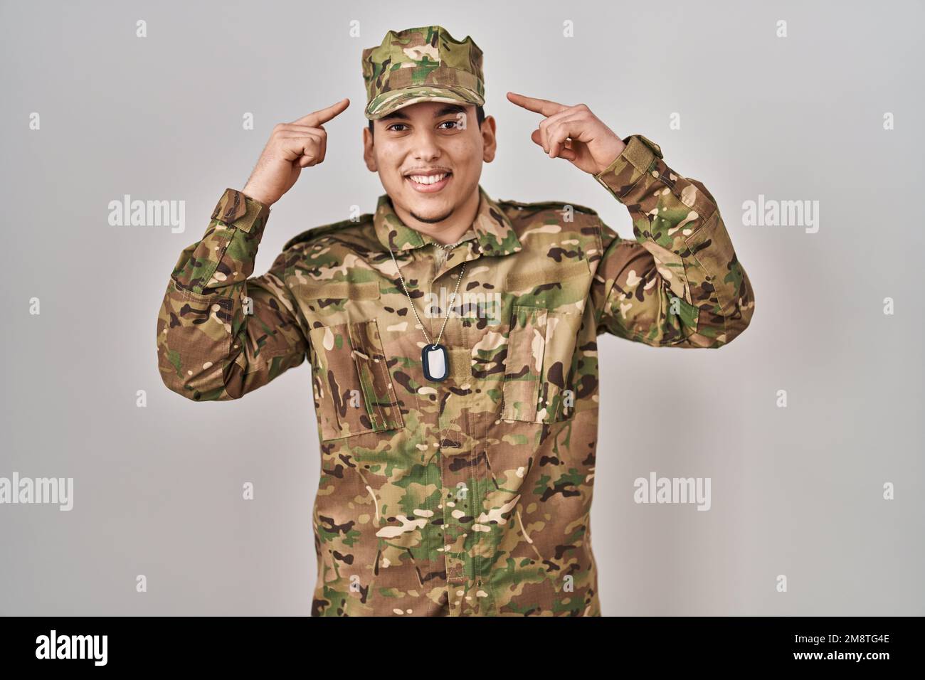 Young arab man wearing camouflage army uniform smiling pointing to head ...