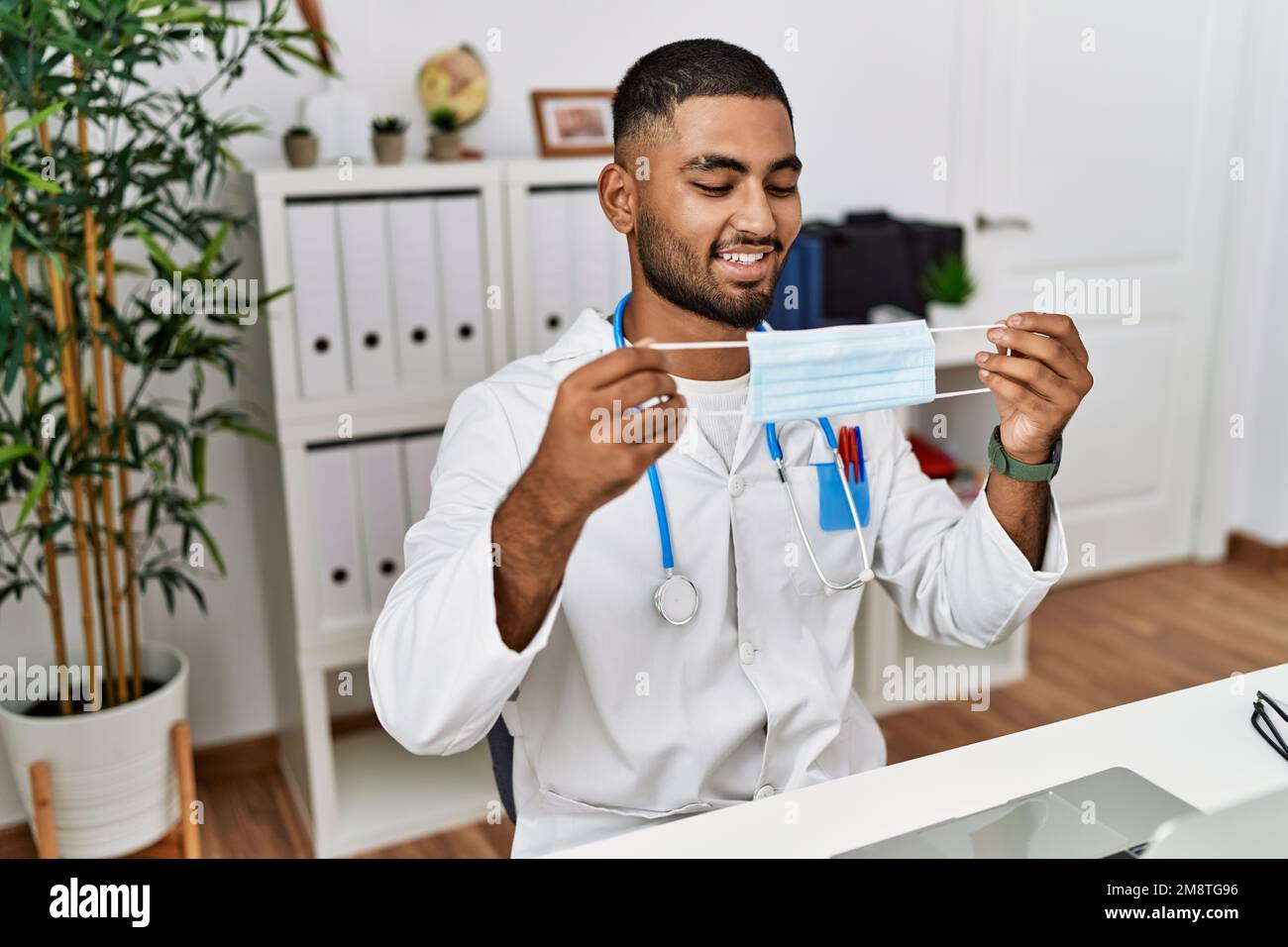Young arab man wearing doctor uniform holding medical mask at clinic ...