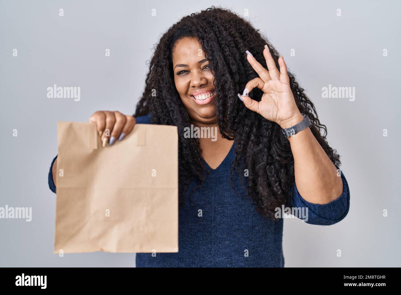 Plus size hispanic woman holding take away paper bag doing ok sign with ...